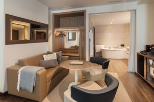 Uma sala de estar moderna com um sofá, poltrona e mesa de centro. Há um espelho na parede, uma área de cozinha ao fundo e um banheiro visível através de uma porta aberta.