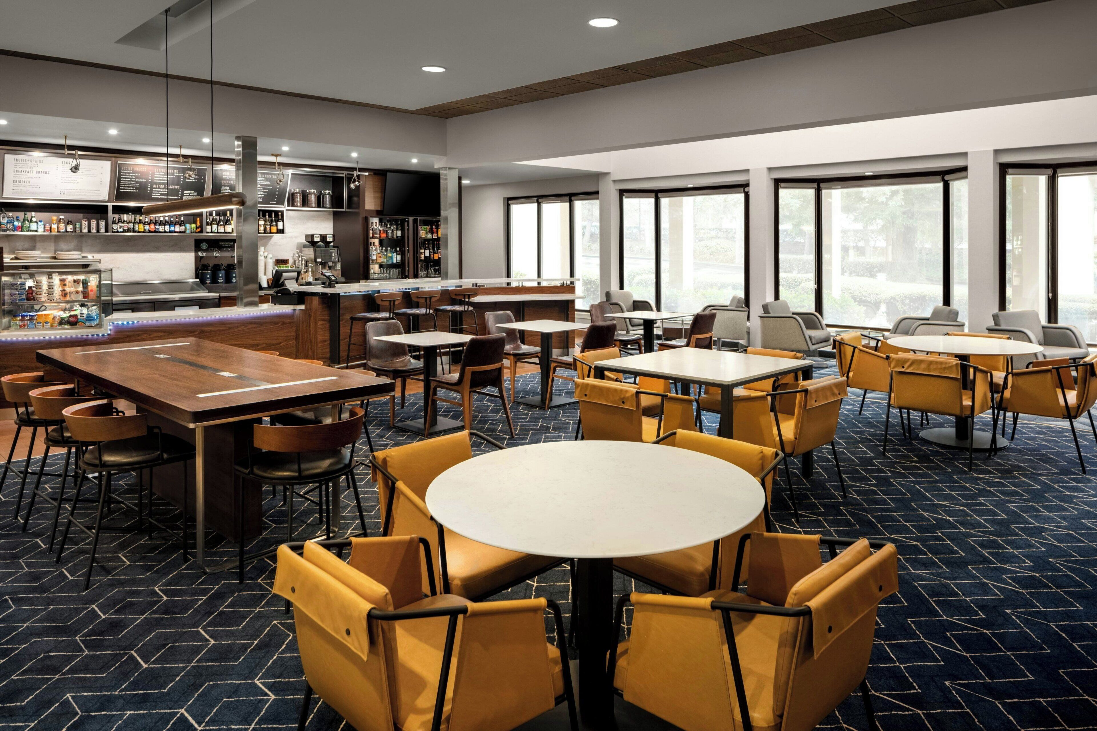 Um restaurante moderno com área de bar, diversas opções de assento, incluindo mesas redondas e cabines, e uma grande janela que oferece vista para o exterior.