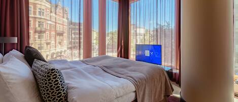 Quarto de hotel com uma cama, televisão e vista para a paisagem urbana através de grandes janelas.