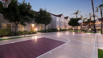 Quadra de basquete ao ar livre com cerca de arame, árvores e casas ao fundo.