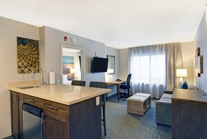 Quarto de hotel com cozinha compacta, área de jantar, TV e espaço de estar com sofá e poltrona.