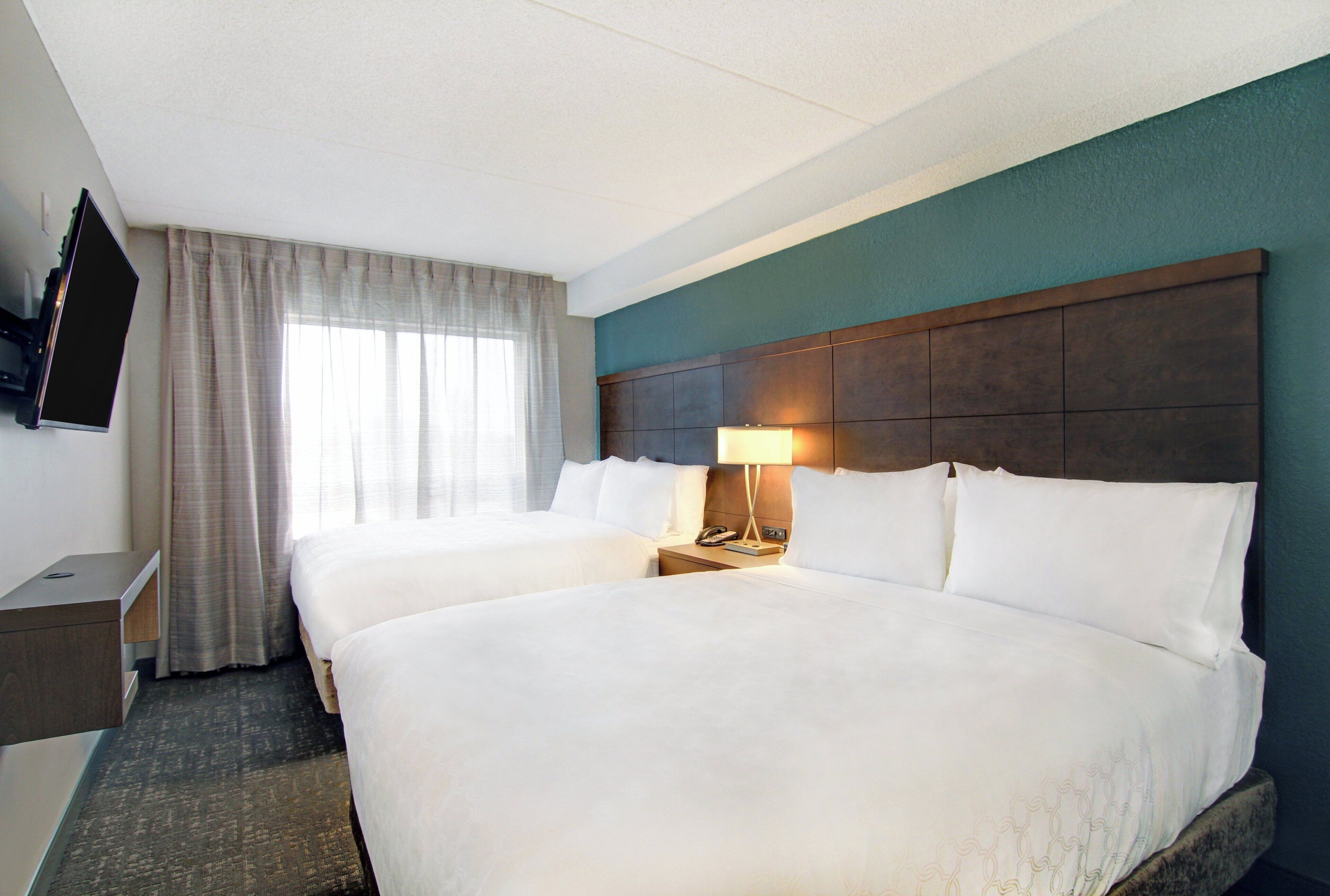 Quarto de hotel com duas camas, televisão na parede, mesa de cabeceira com abajur e janela com cortinas.