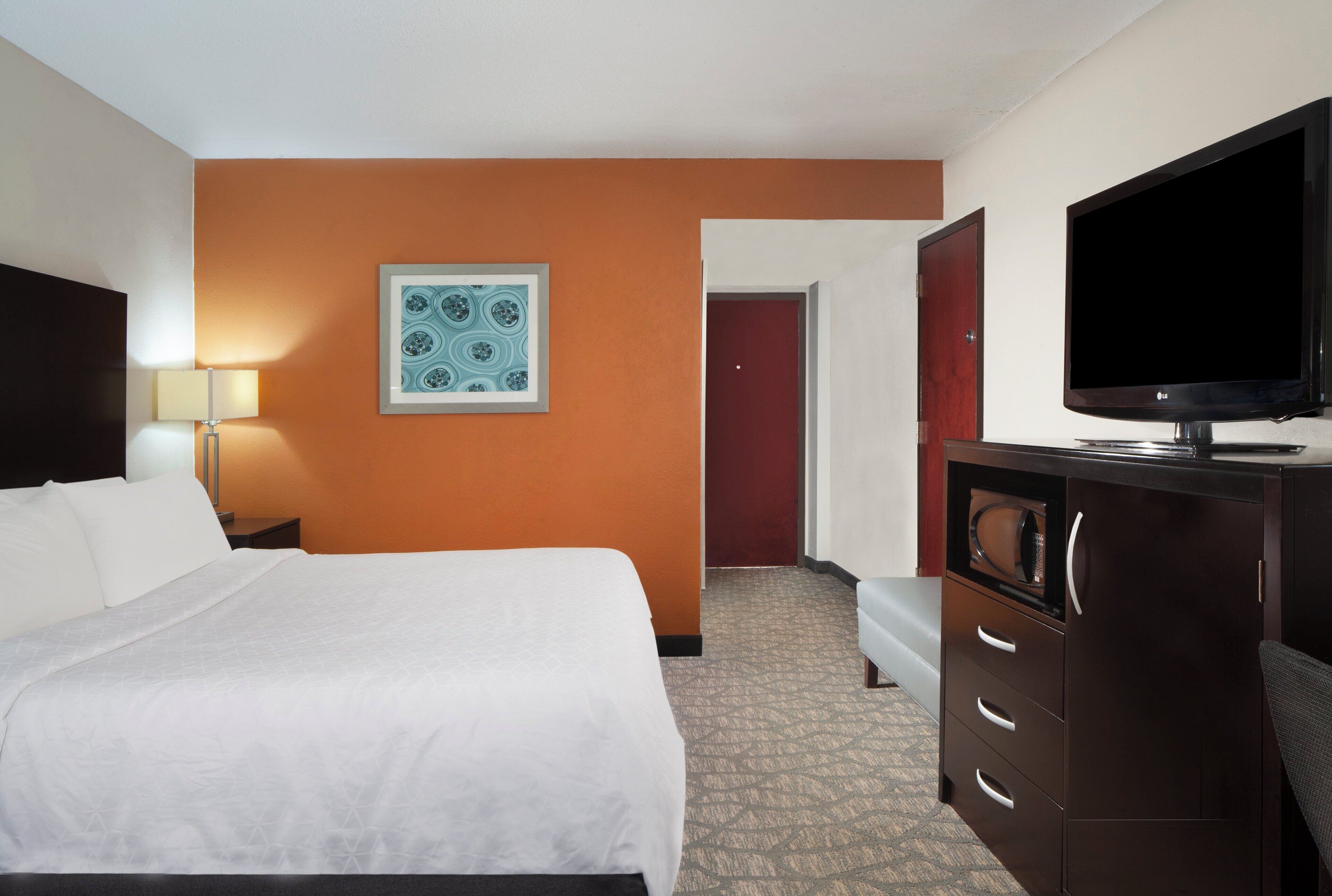 Quarto de hotel com uma cama, penteadeira, televisão, luminária e um quadro na parede.