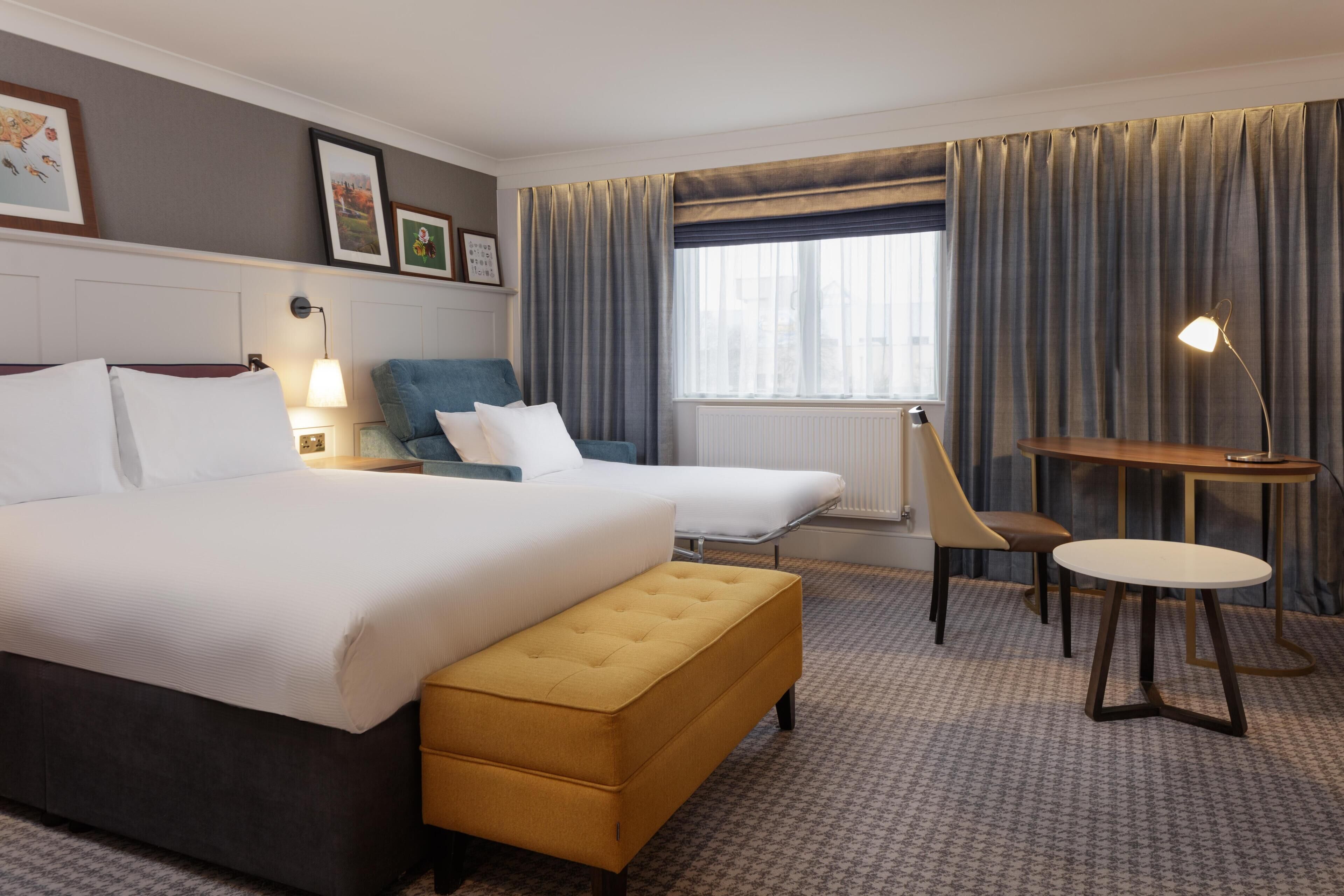 Quarto de hotel com uma cama grande, um sofá, uma mesa pequena, uma cadeira e uma janela com cortinas.