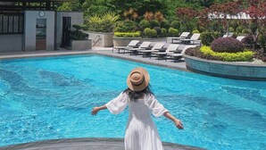 Uma área de piscina com uma pessoa de vestido e chapéu branco, cercada por espreguiçadeiras e um edifício de design moderno.