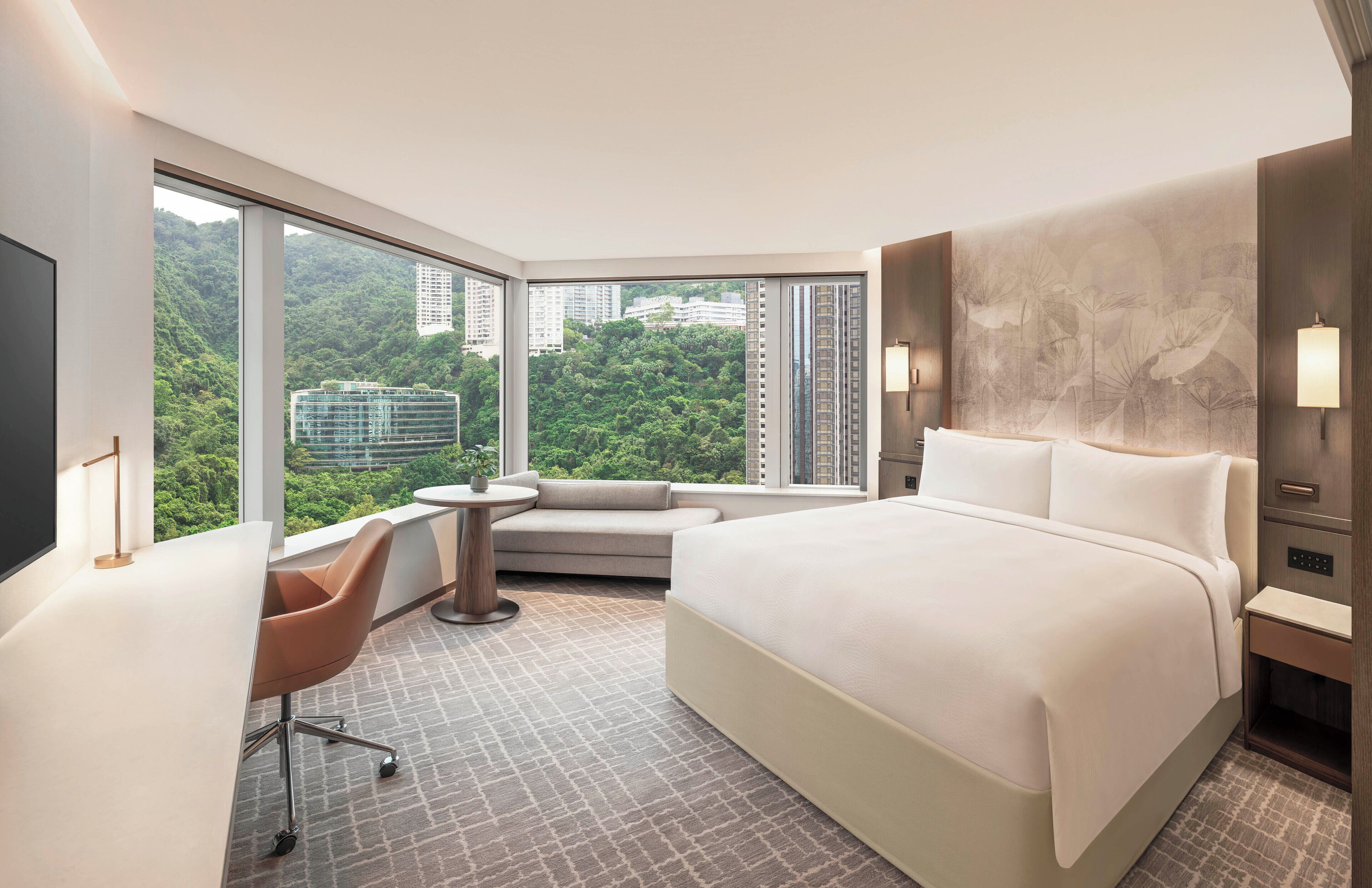 Quarto de hotel moderno com uma cama grande, uma mesa de escritório, uma cadeira e uma pequena mesa. Há uma janela ampla com vista para o horizonte urbano e áreas verdes.