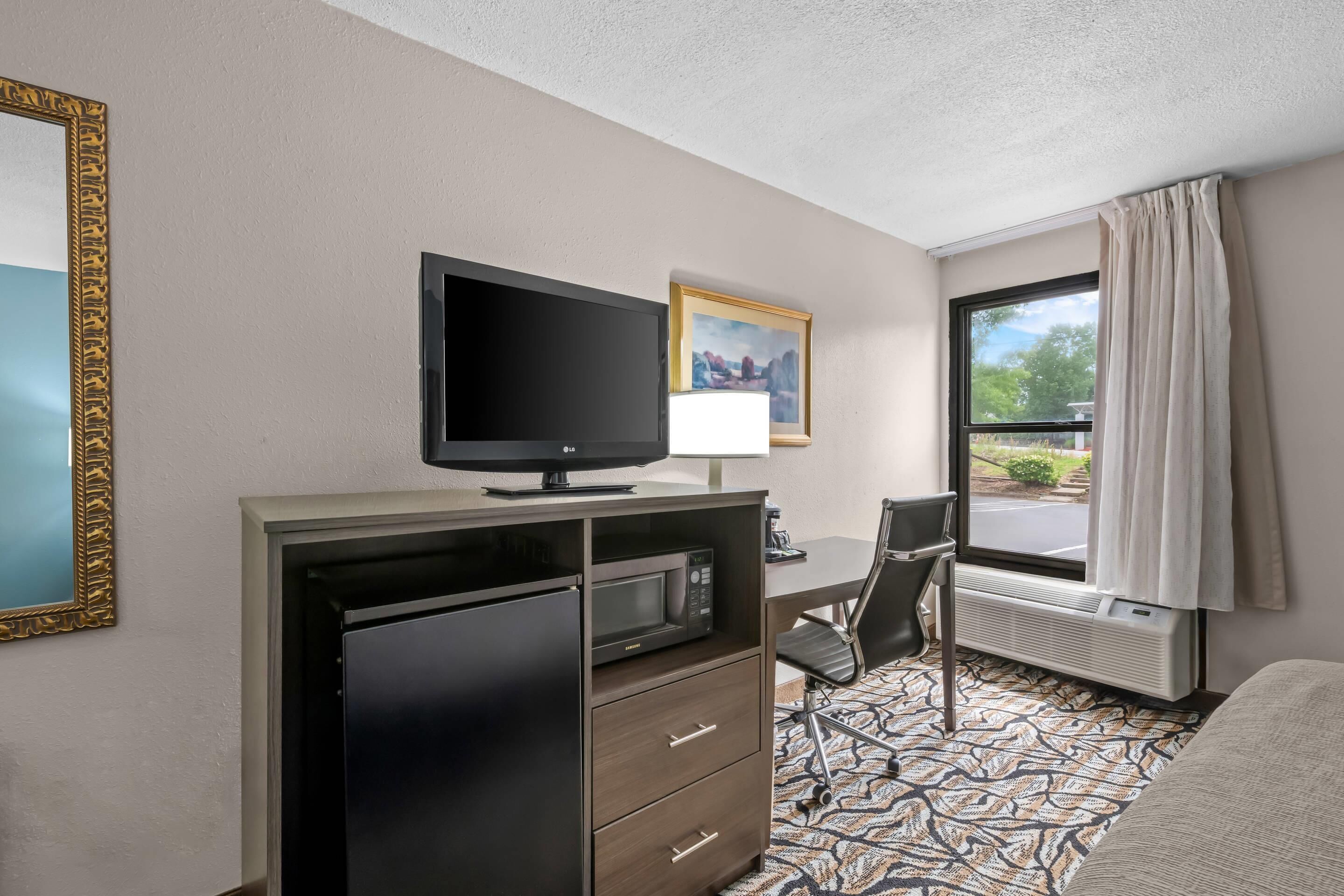 Quarto de hotel com televisão de tela plana, escrivaninha com cadeira, cama, janela com cortinas e carpete estampado.