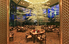 Uma ampla área de jantar com várias mesas e cadeiras, uma árvore ornamental central e um design arquitetônico moderno com grandes janelas.