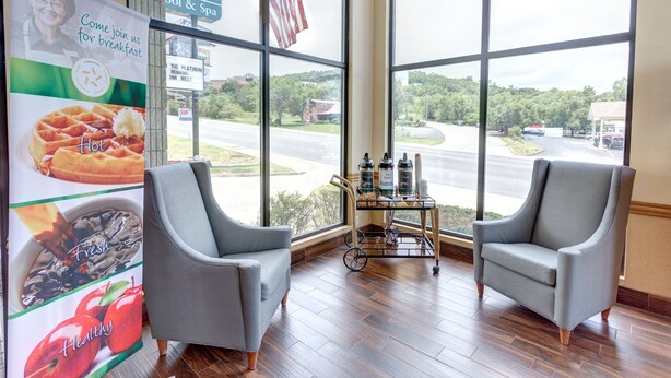 Um saguão de hotel com dois poltronas cinzentas, um carrinho de café e uma grande janela com vista para uma rua e vegetação.