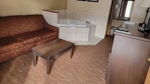 Quarto de hotel com um sofá marrom, uma mesa de centro de madeira, uma banheira branca e uma pequena escrivaninha com abajur.
