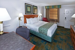 Quarto de hotel com cama, duas luminárias de cabeceira, uma cadeira, escrivaninha e porta.
