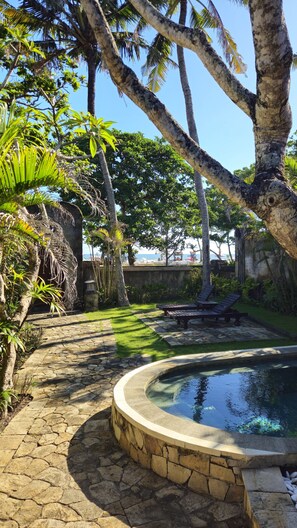 Uma área pavimentada com pedras, com piscina, rodeada por palmeiras e vegetação.