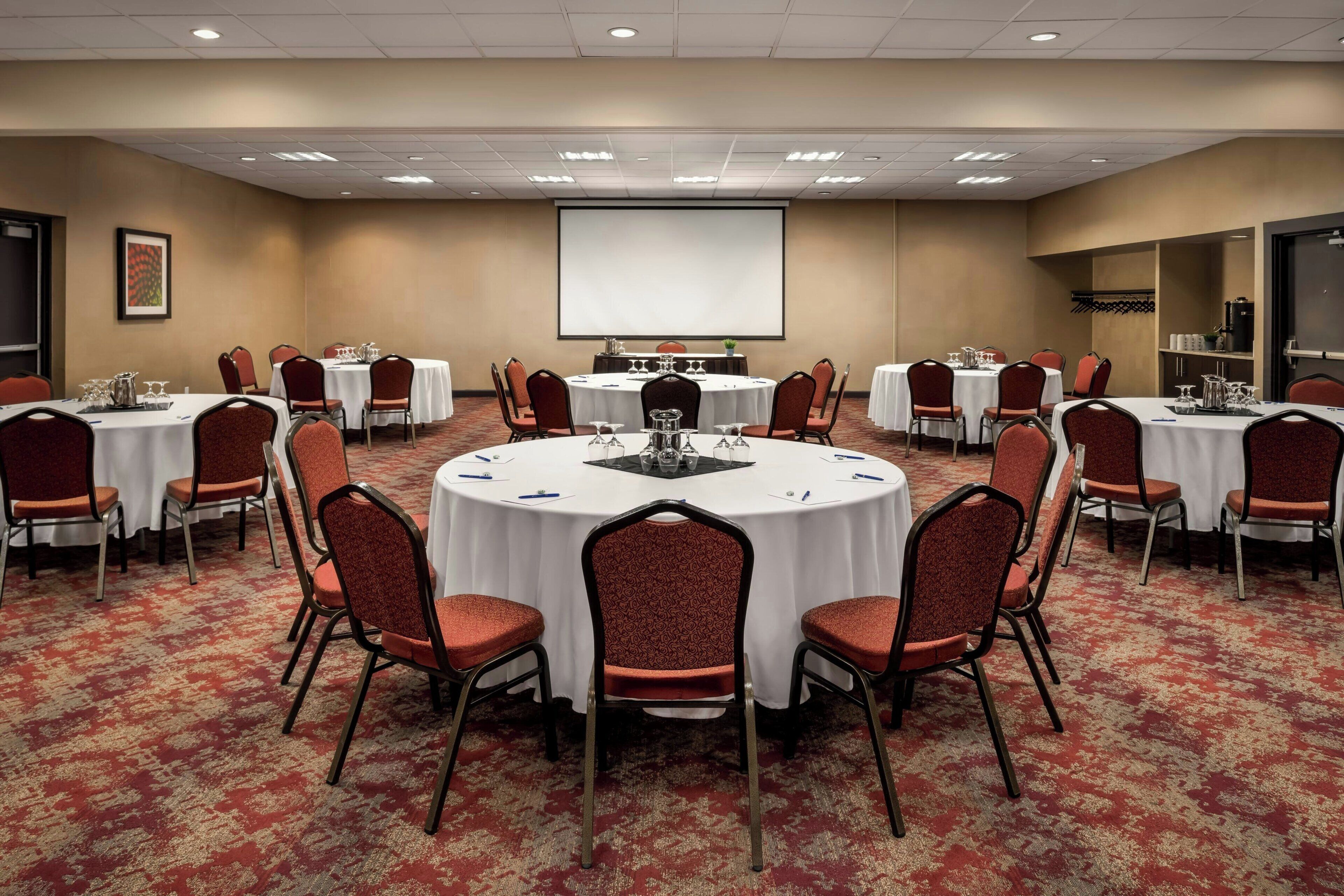 Sala de conferências com mesas redondas, cadeiras vermelhas, toalhas de mesa brancas e uma tela de projetor.