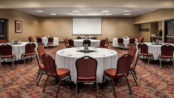 Sala de conferências com mesas redondas, cadeiras vermelhas, toalhas de mesa brancas e uma tela de projetor.