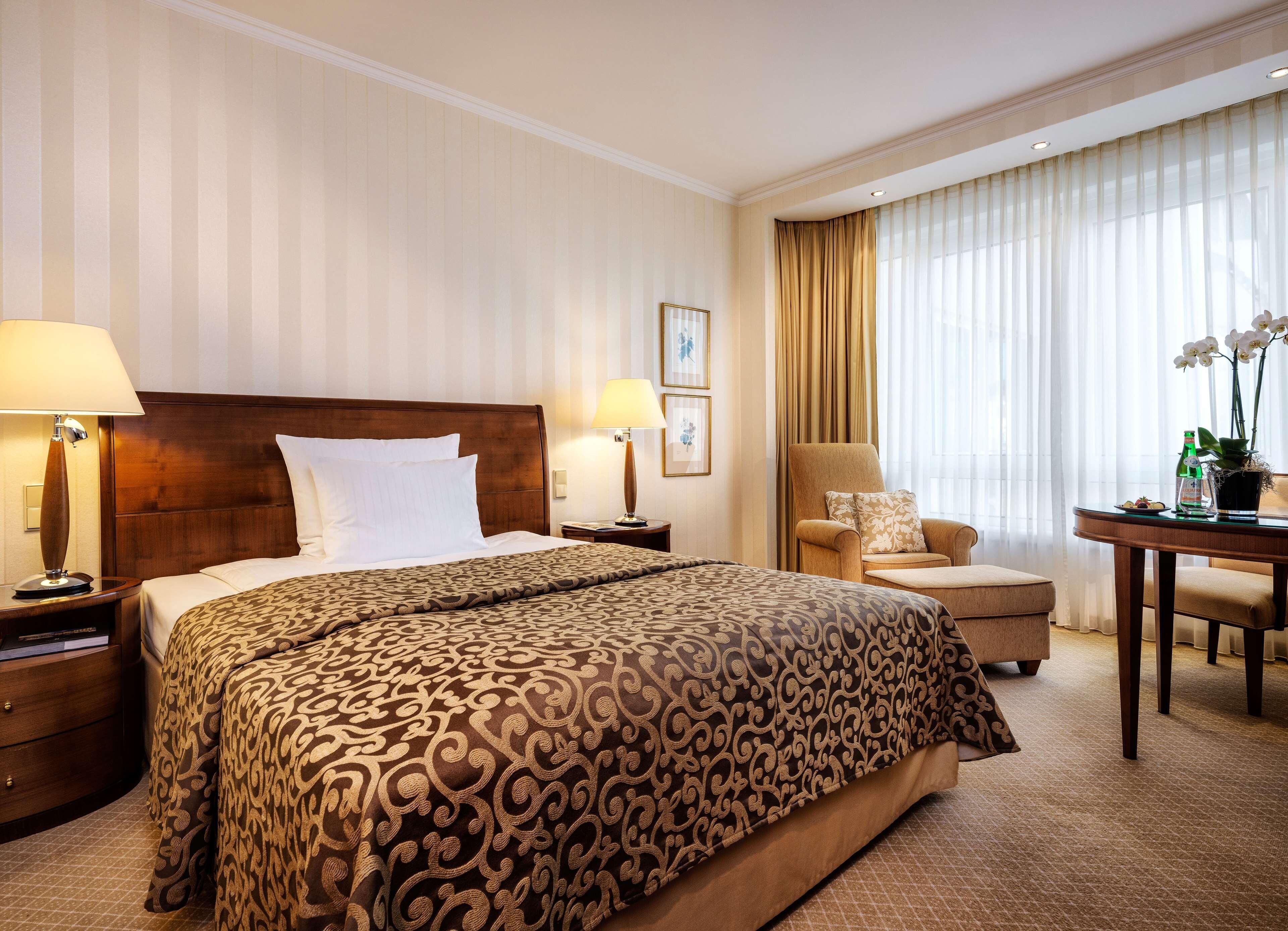 Quarto de hotel com uma cama grande, duas mesas de cabeceira com abajures, uma cadeira, uma pequena mesa com uma planta e uma janela com cortinas.