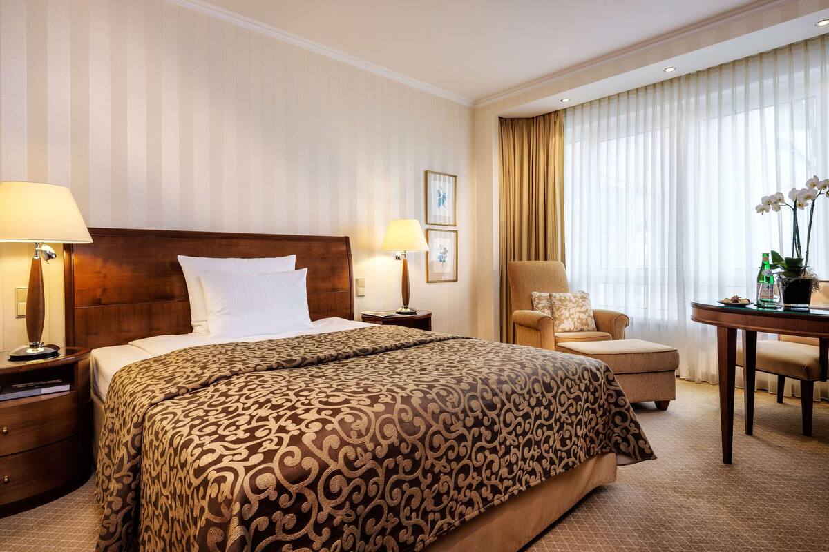 Quarto de hotel com uma cama grande, duas mesas de cabeceira com abajures, uma cadeira, uma pequena mesa com uma planta e uma janela com cortinas.
