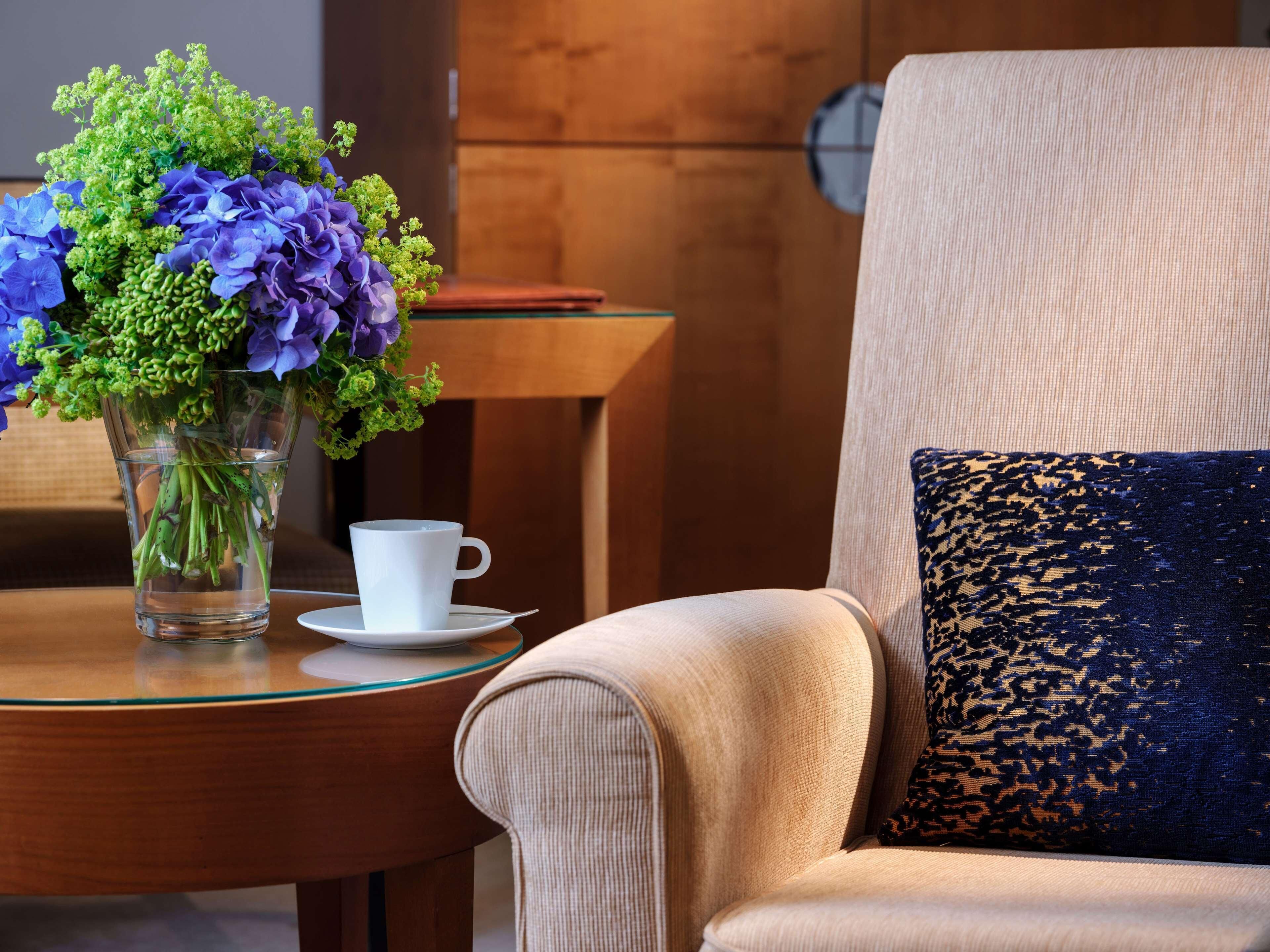 Um quarto com uma mesa de vidro redonda, uma xícara e pires brancos, uma poltrona bege com almofada estampada e um vaso com flores verdes e roxas.