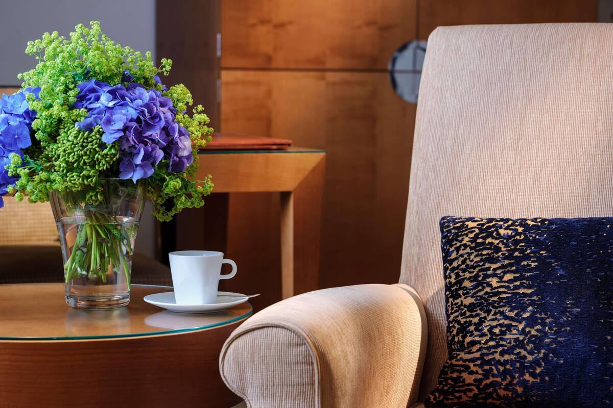 Um quarto com uma mesa de vidro redonda, uma xícara e pires brancos, uma poltrona bege com almofada estampada e um vaso com flores verdes e roxas.