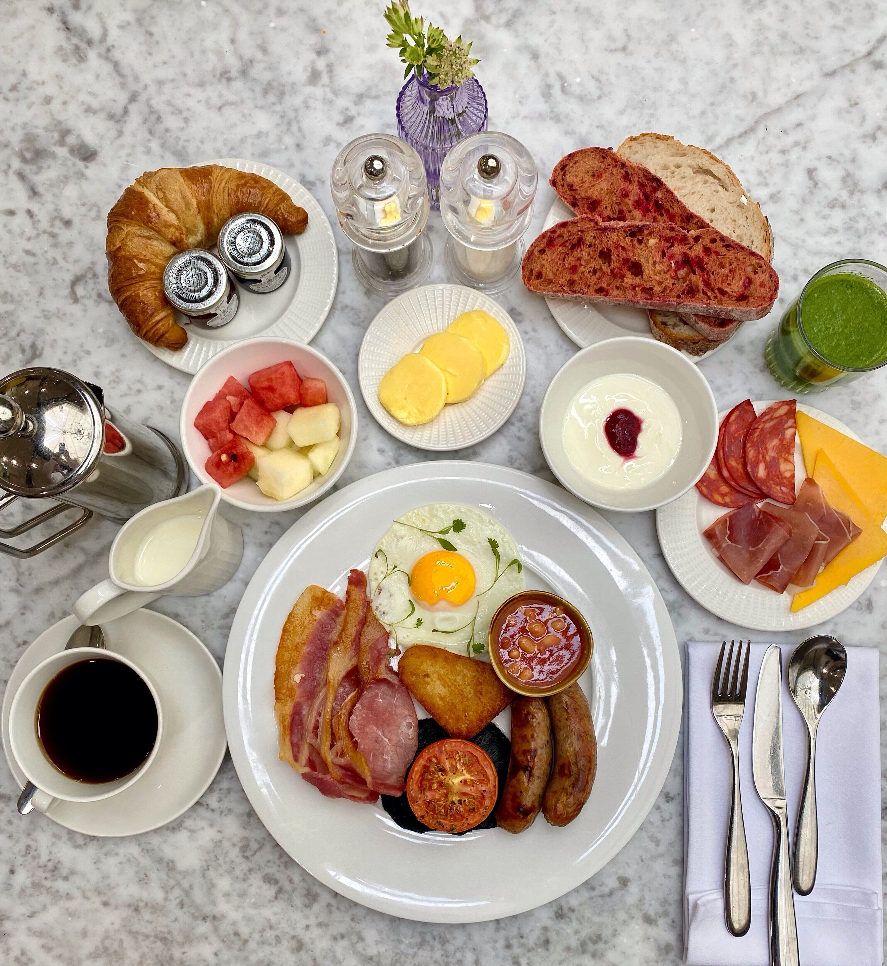 Uma mesa de mármore com uma variedade de alimentos para o café da manhã, incluindo croissants, torradas, ovos, bacon, linguiça e frutas e queijos diversos.
