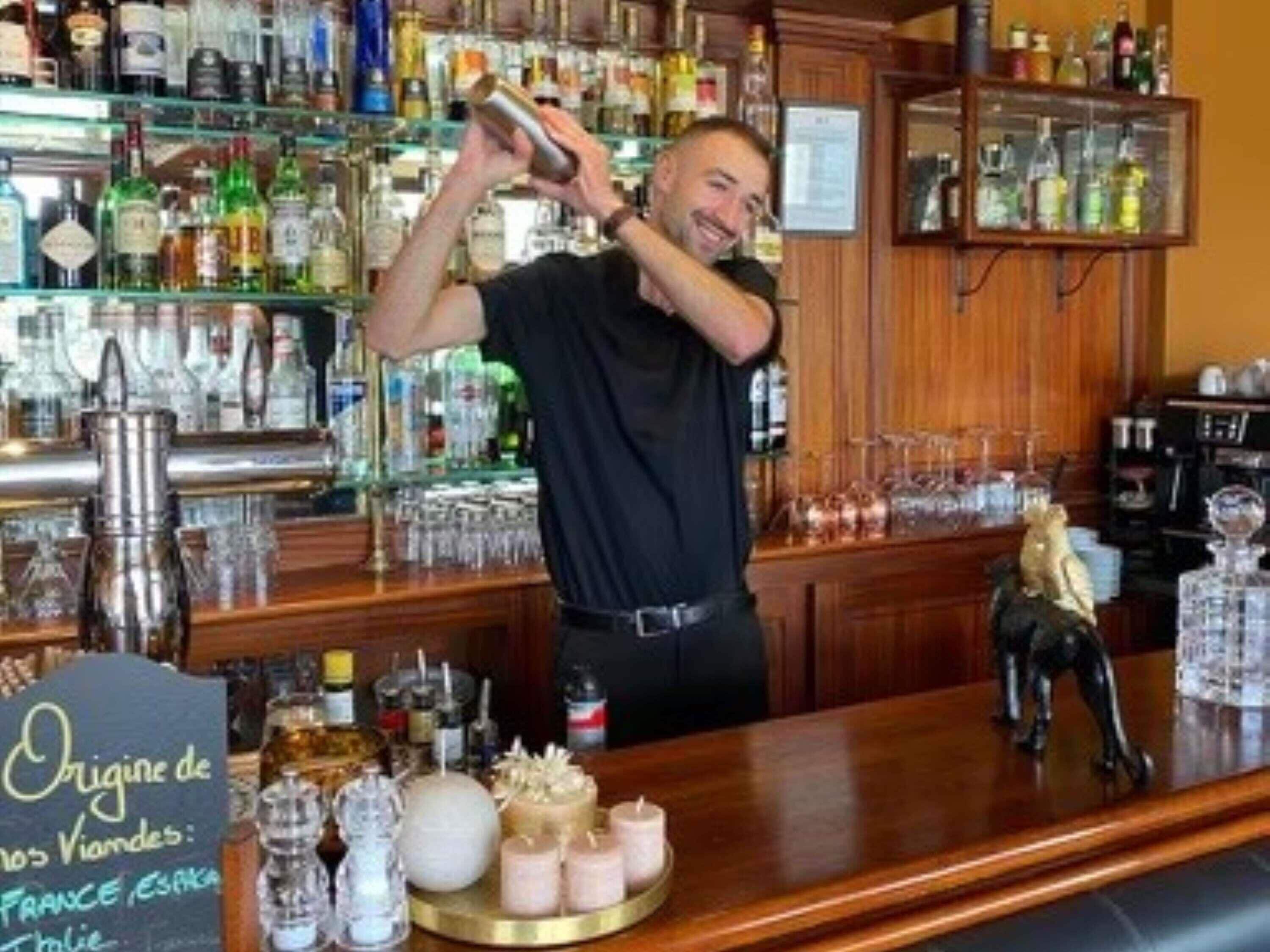 Bar com garçom, prateleiras cheias de garrafas de bebidas e cardápio em lousa.