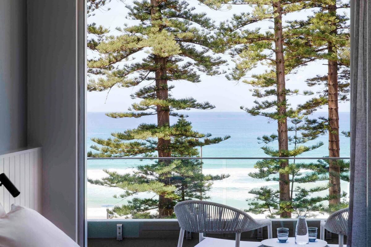Quarto de hotel com uma janela ampla com vista para uma praia e altas pinhas. Há uma cama, uma mesa pequena com cadeiras e vista para o oceano.