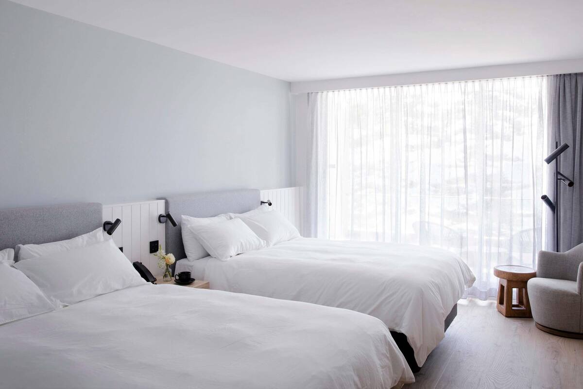 Quarto de hotel com duas camas, um banquinho de madeira, uma cadeira e uma janela grande com cortinas finas.