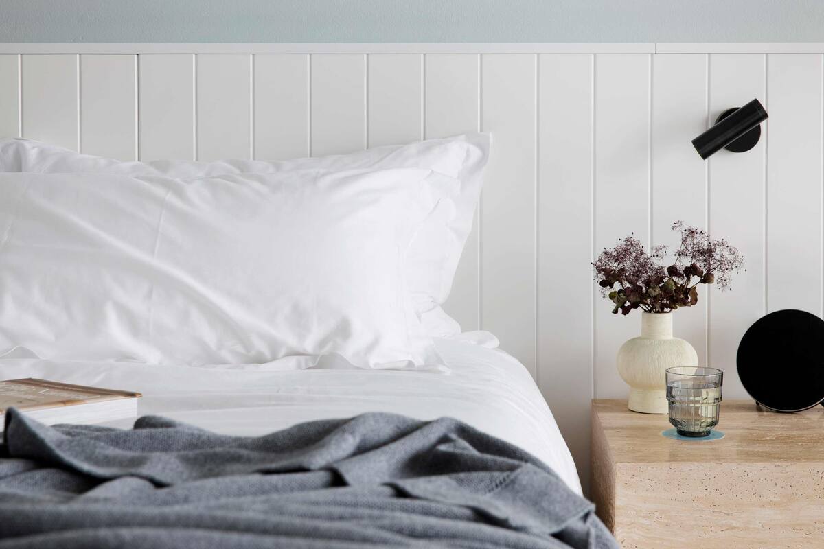 Cama bem arrumada com roupa de cama branca, manta cinza, mesa de cabeceira com um vaso de flores, um copo de água e um objeto preto na parede.