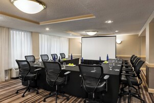 Uma sala de conferências com uma mesa comprida, cadeiras pretas e uma tela de projetor.