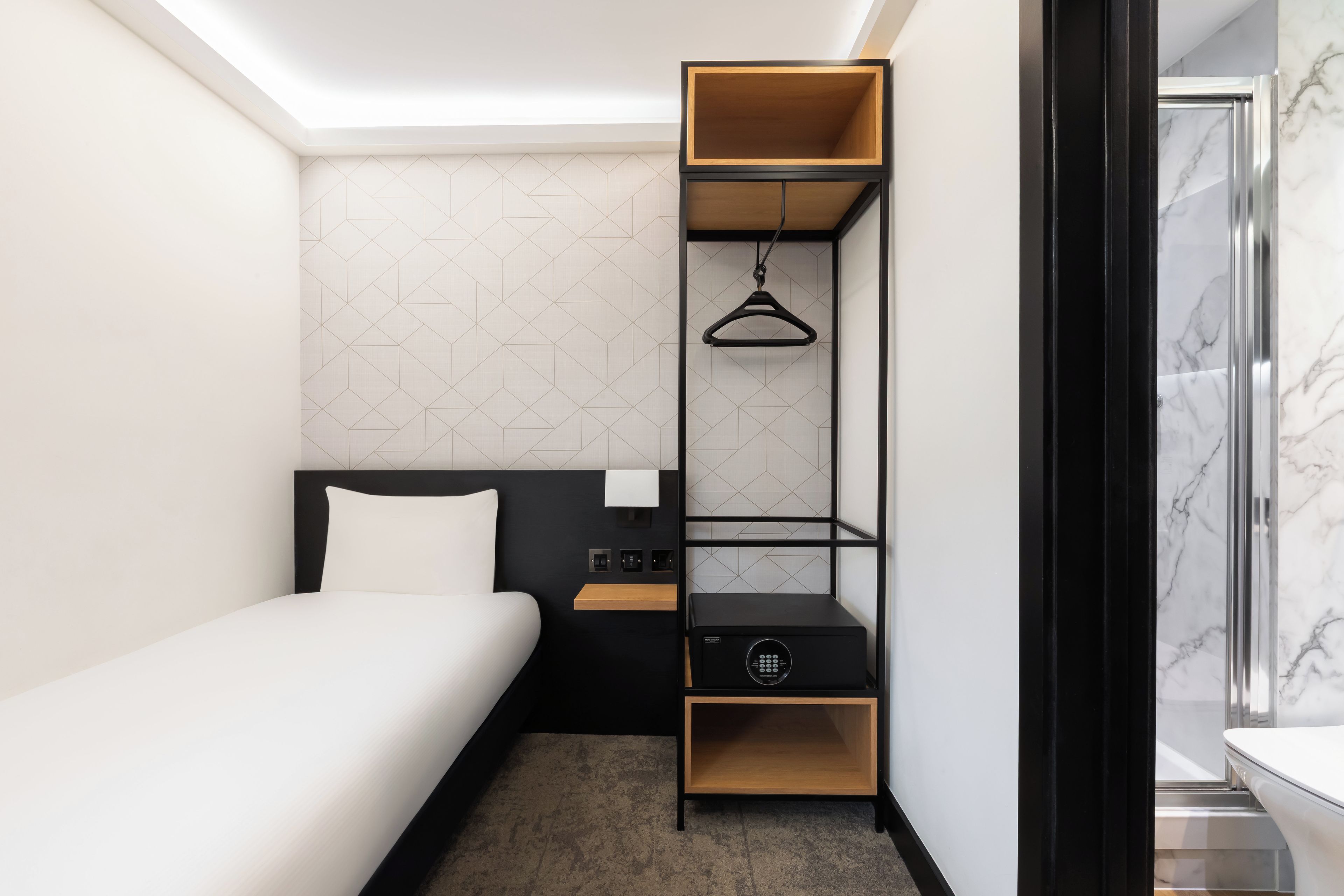 Quarto de hotel compacto com cama, mesa de cabeceira pequena, guarda-roupa com espaço para pendurar e área de chuveiro.