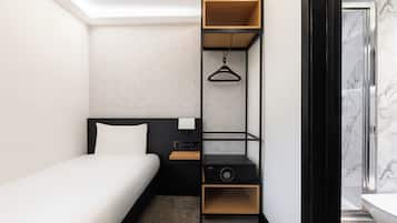 Quarto de hotel compacto com cama, mesa de cabeceira pequena, guarda-roupa com espaço para pendurar e área de chuveiro.