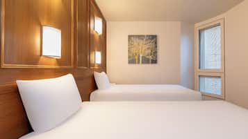 Quarto de hotel com duas camas, cabeceira de madeira, luminárias fixadas na parede, um quadro e uma janela com persianas.