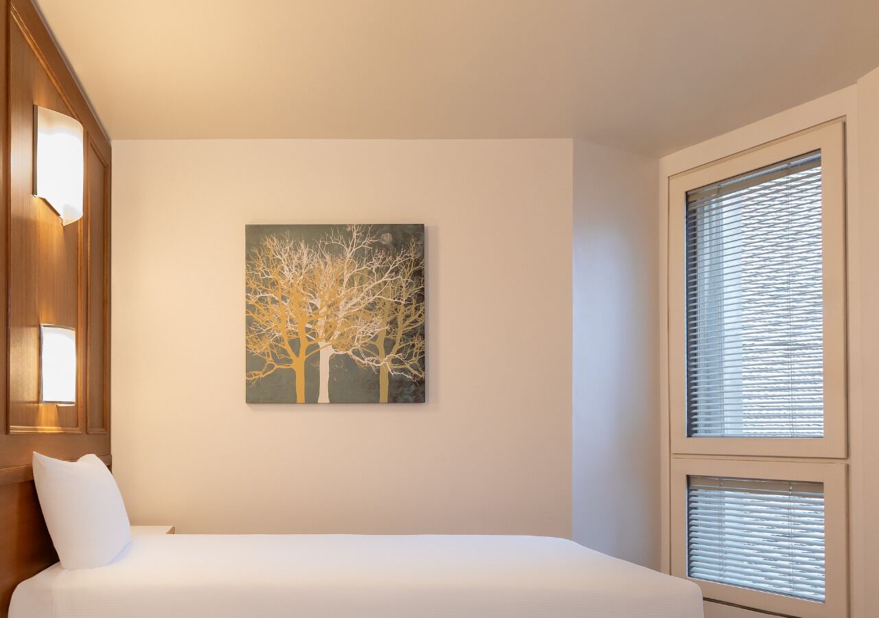 Quarto de hotel com uma cama, um quadro de árvores na parede e uma janela com persianas.