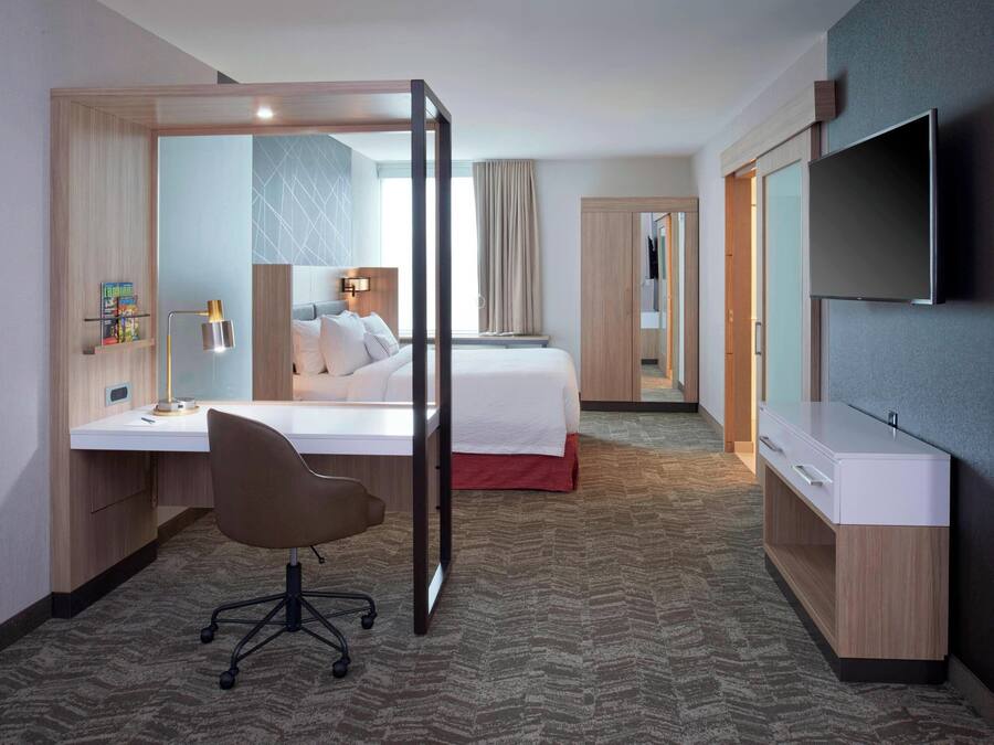 Quarto de hotel com cama, escrivaninha, cadeira, televisão e espelho.