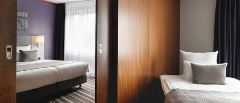 Quarto de hotel com duas camas, guarda-roupa de madeira, tapete listrado e uma janela grande com cortinas.