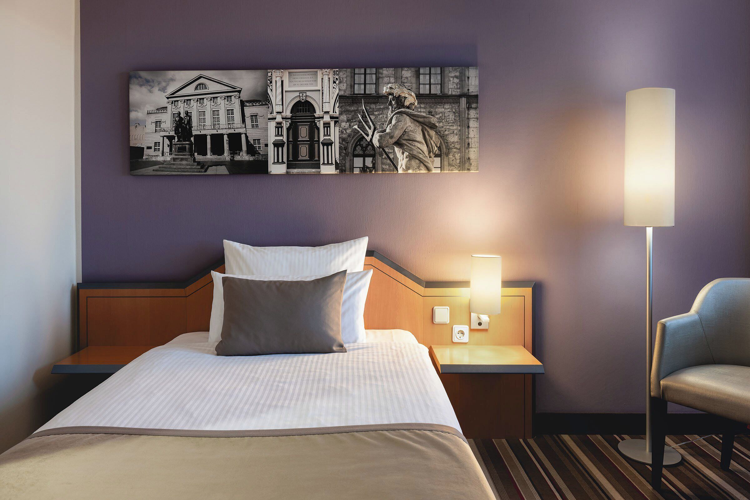Quarto de hotel com cama, mesa de cabeceira, luminária, cadeira e uma obra de arte na parede retratando um edifício e uma estátua.