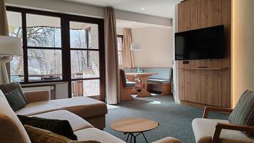 Uma sala de estar moderna com sofá, poltrona e uma mesa pequena. Uma TV está montada na parede, e há uma janela com cortinas.