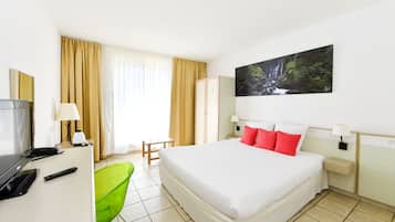 Quarto de hotel com uma cama, uma escrivaninha com um computador, uma cadeira verde, uma televisão e um quadro de uma cascata.
