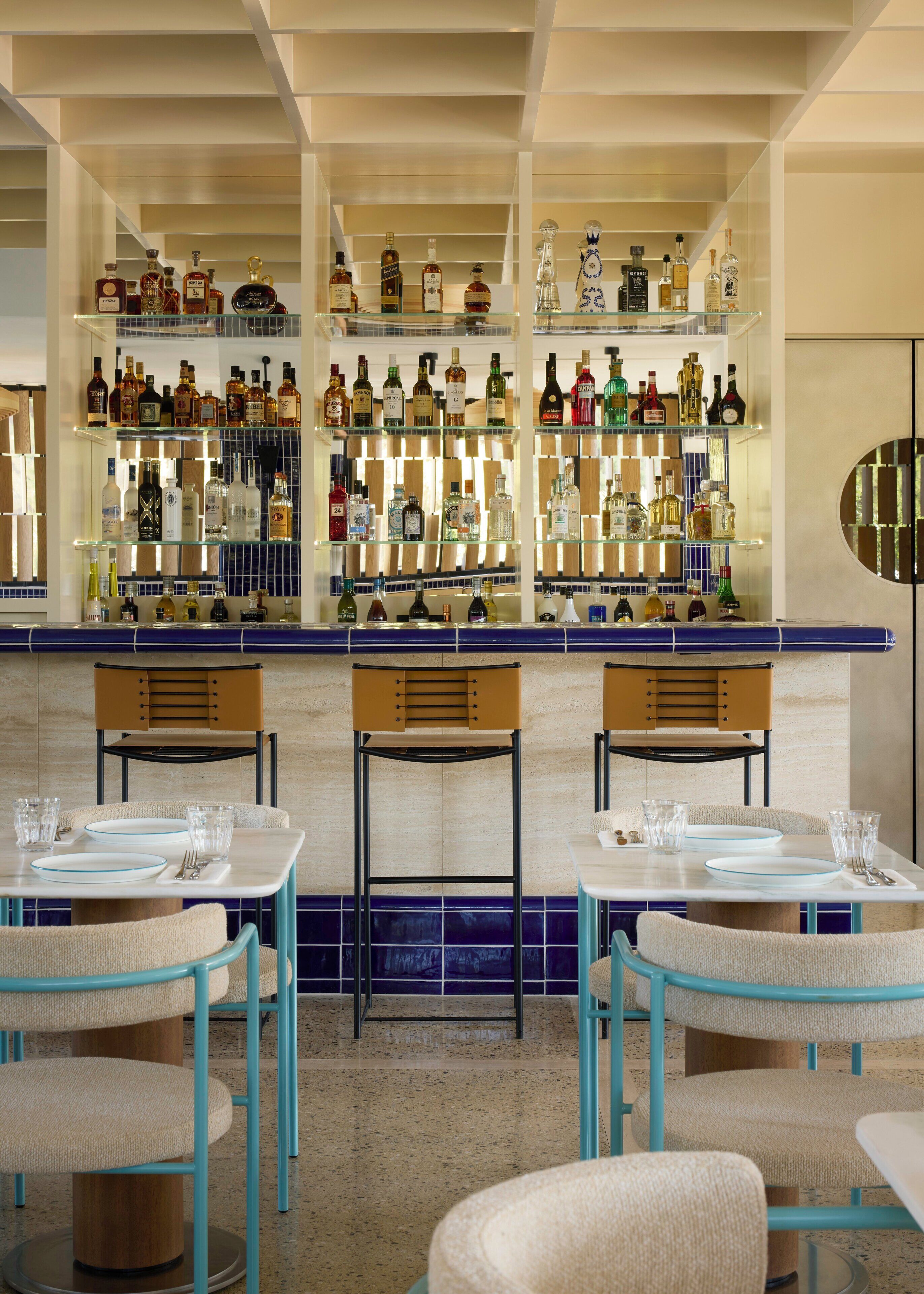Uma área de bar com várias prateleiras de garrafas de bebidas, banquetas e mesas preparadas para refeições.