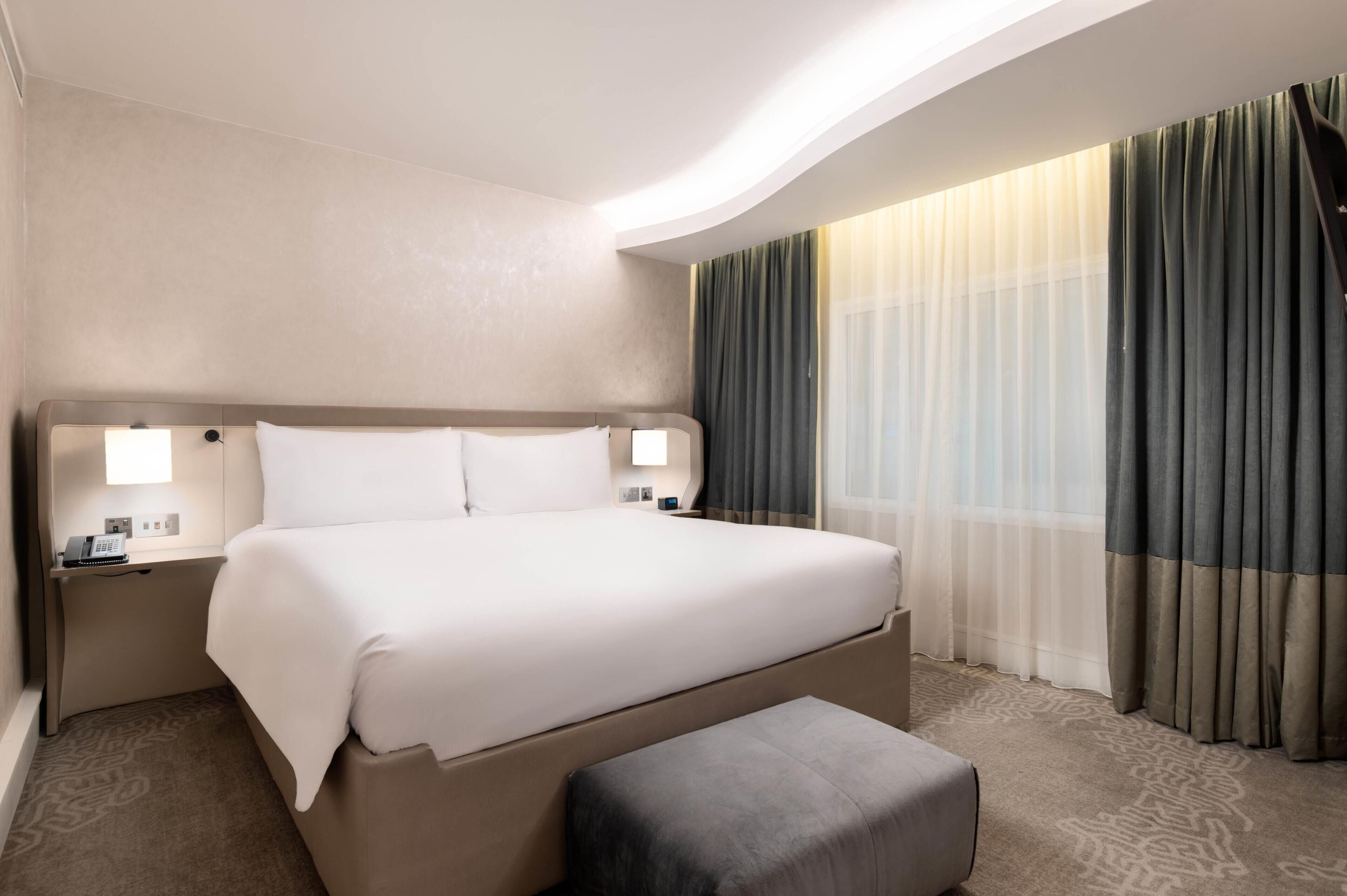 Quarto de hotel com uma cama grande, mesas de cabeceira, luminária, telefone e janela com cortinas.