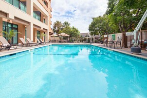 Piscina com espreguiçadeiras e guarda-sóis, rodeada por um edifício com vários andares e janelas.
