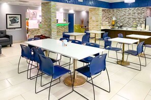 Uma sala de jantar moderna com mesas brancas e cadeiras azuis, parede de destaque em pedra e um balcão com máquina de café.