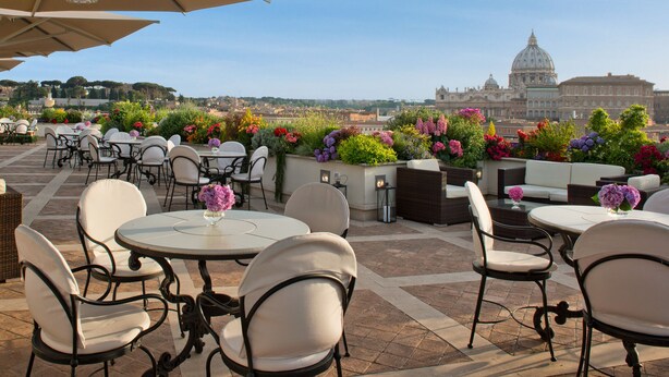 Terraço ao ar livre com mesas e cadeiras, rodeado por plantas e flores em vasos, com vista para um edifício histórico.