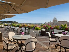 Terraço ao ar livre com mesas e cadeiras, rodeado por plantas e flores em vasos, com vista para um edifício histórico.