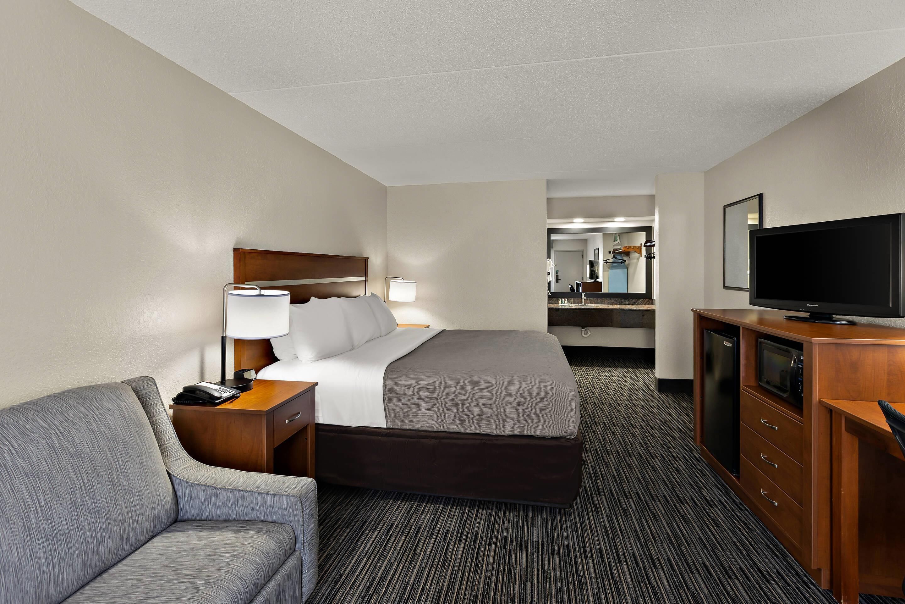 Quarto de hotel com cama, escrivaninha com luminária, televisão e área de estar.