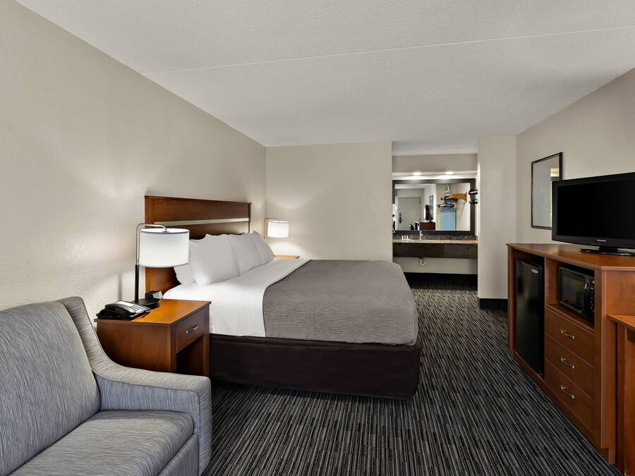 Quarto de hotel com cama, escrivaninha com luminária, televisão e área de estar.
