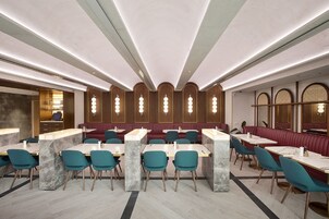 Restaurante moderno com mesas de bancada em mármore e cadeiras cor de azul-turquesa, apresentando uma área de estar central com assentos acolchoados vermelhos e iluminação linear no teto.