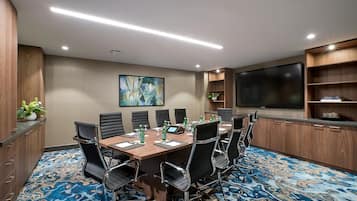 Uma sala de conferências com uma mesa grande, cadeiras pretas, uma TV de tela plana e um quadro decorativo na parede.