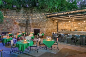 Área de refeições ao ar livre com paredes de pedra, luzes de corda e um bar com garrafas. Mesas montadas com cadeiras e velas.