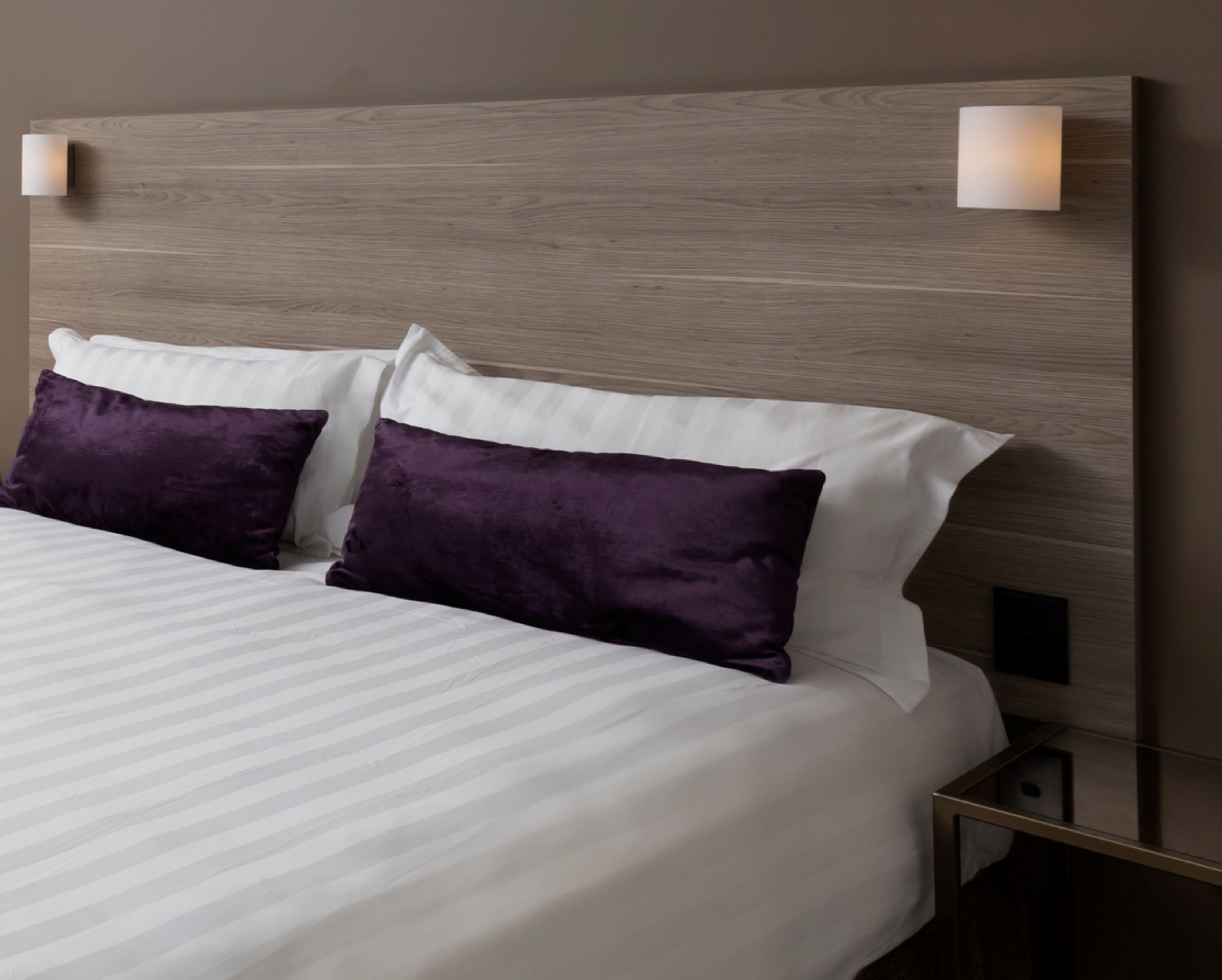 Cama bem arrumada com almofadas brancas e roxas, cabeceira de madeira e duas luminárias fixadas na parede.