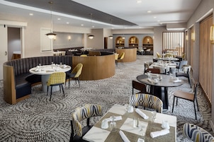 Uma área de refeições de hotel com mesas redondas, assentos acolchoados e um tapete estampado.